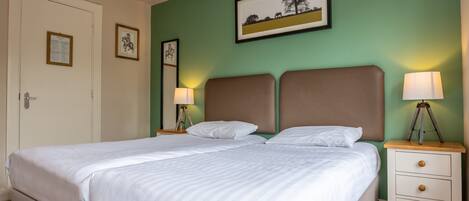 Uma cama de casal com lençóis brancos, uma mesa de cabeceira de madeira com abajur, e uma parede verde com obras de arte emolduradas.
