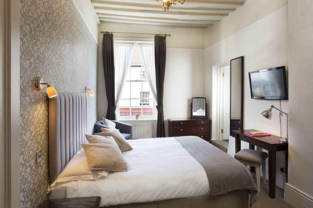 Quarto de hotel com uma cama, uma escrivaninha com computador, televisão e janela com cortinas.