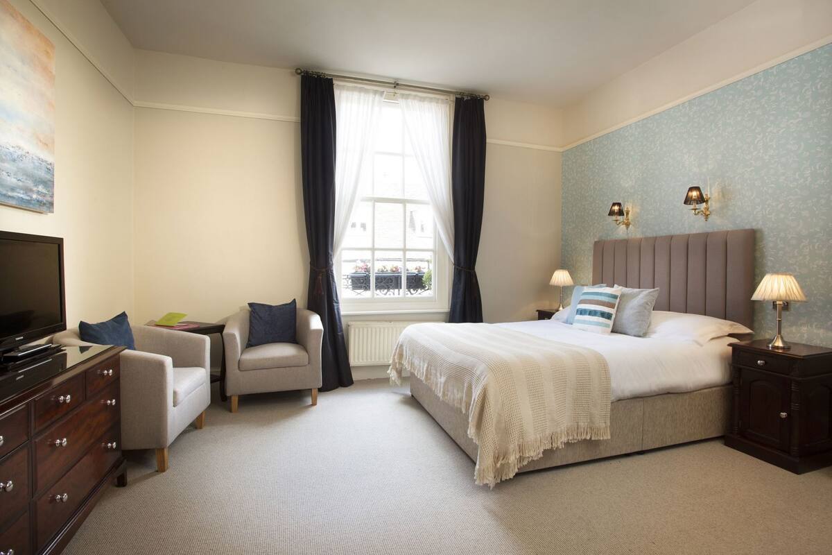 Quarto de hotel com cama, televisão, cadeira, penteadeira e janela com cortinas.