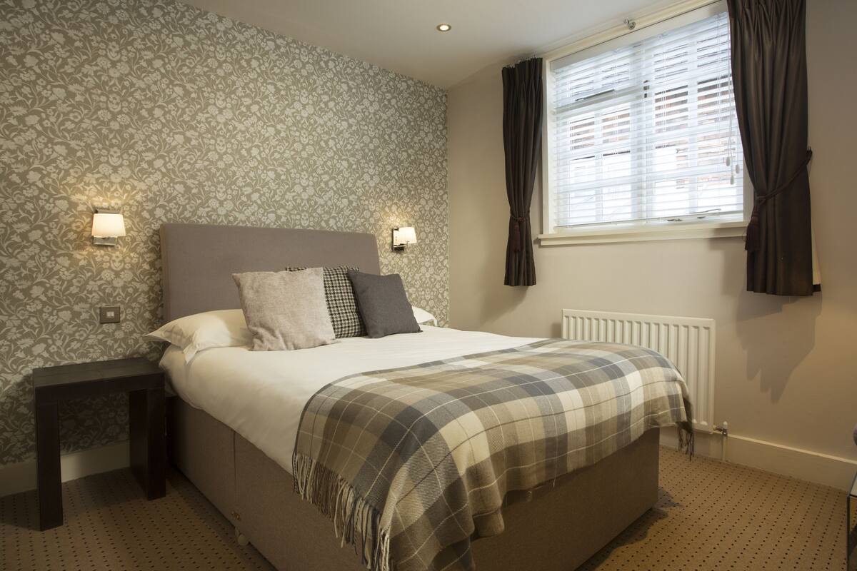 Um quarto com cama, duas luminárias de cabeceira, janela com cortinas e um radiador.