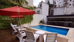 Área de piscina com espreguiçadeiras brancas e um guarda-sol vermelho, cercada por um deck de madeira e um edifício branco com vegetação.