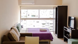 Uma sala de estar moderna com uma mesa de jantar de vidro, um sofá, uma televisão e uma grande janela com vista para a cidade.