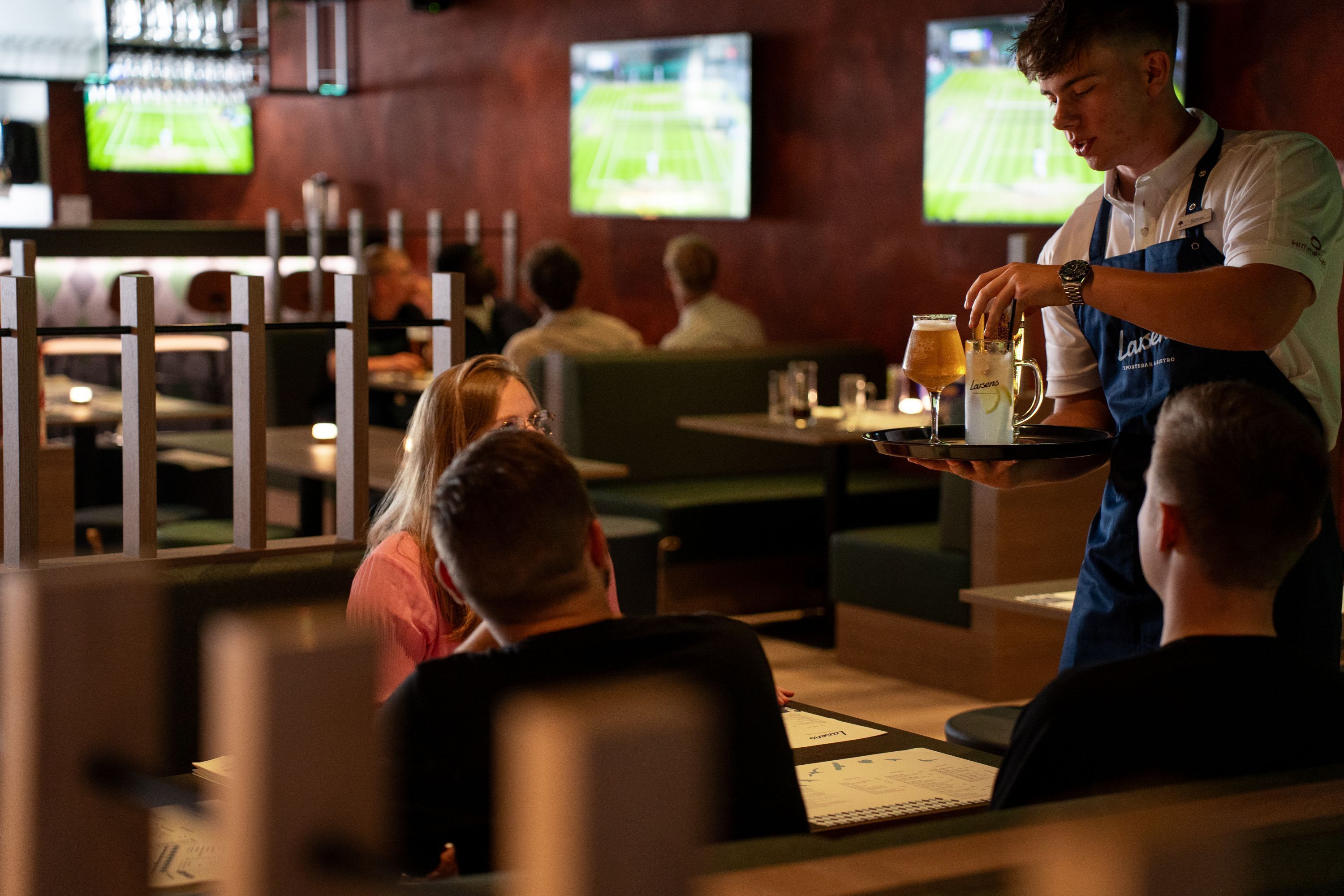 Um restaurante com mesas e cadeiras, um garçom servindo bebidas e uma TV exibindo uma partida de tênis.