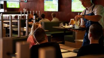 Um restaurante com mesas e cadeiras, um garçom servindo bebidas e uma TV exibindo uma partida de tênis.