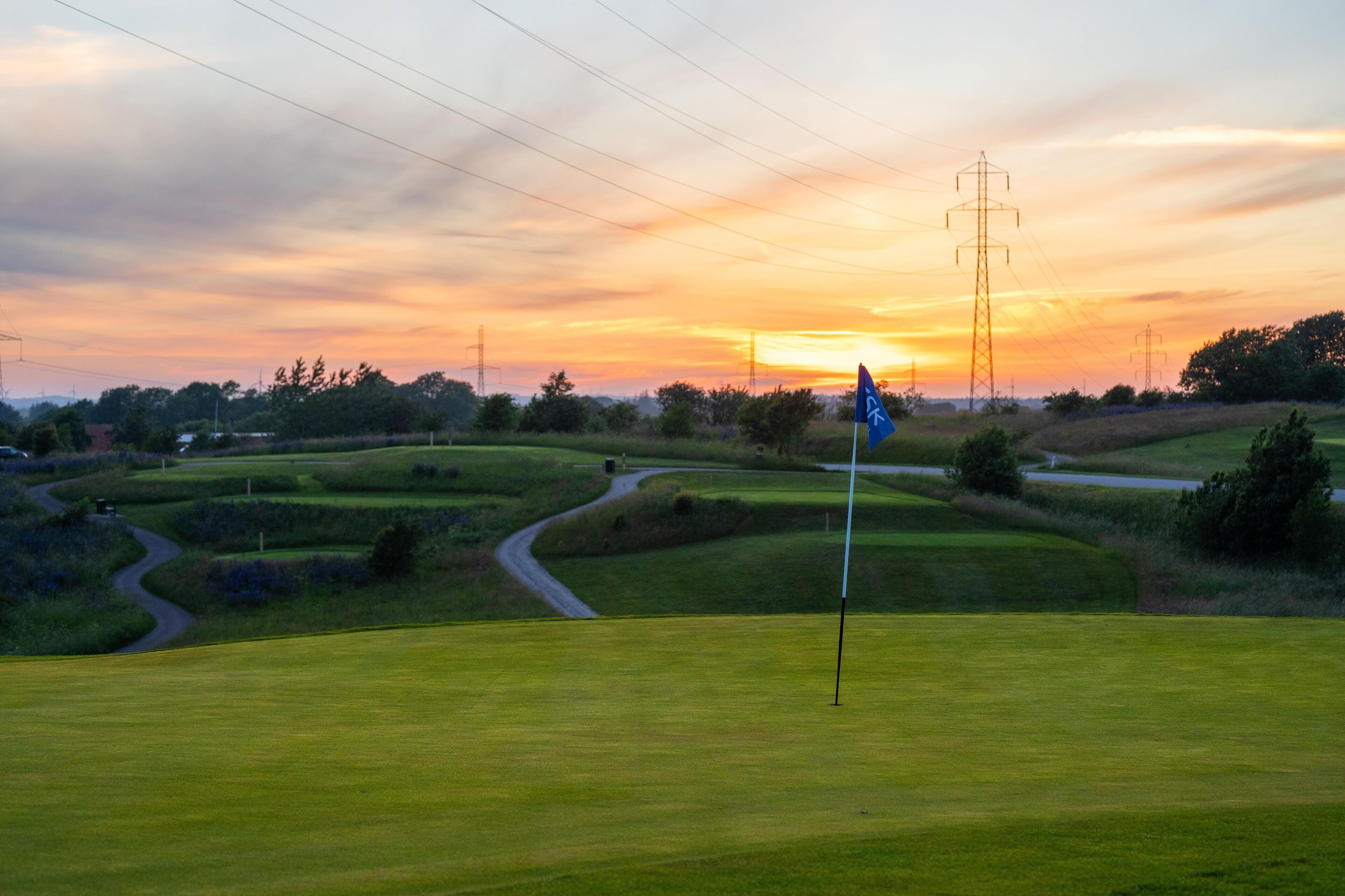 Um campo de golfe ao pôr do sol, com um mastro de bandeira em primeiro plano, linhas de eletricidade e um caminho sinuoso.