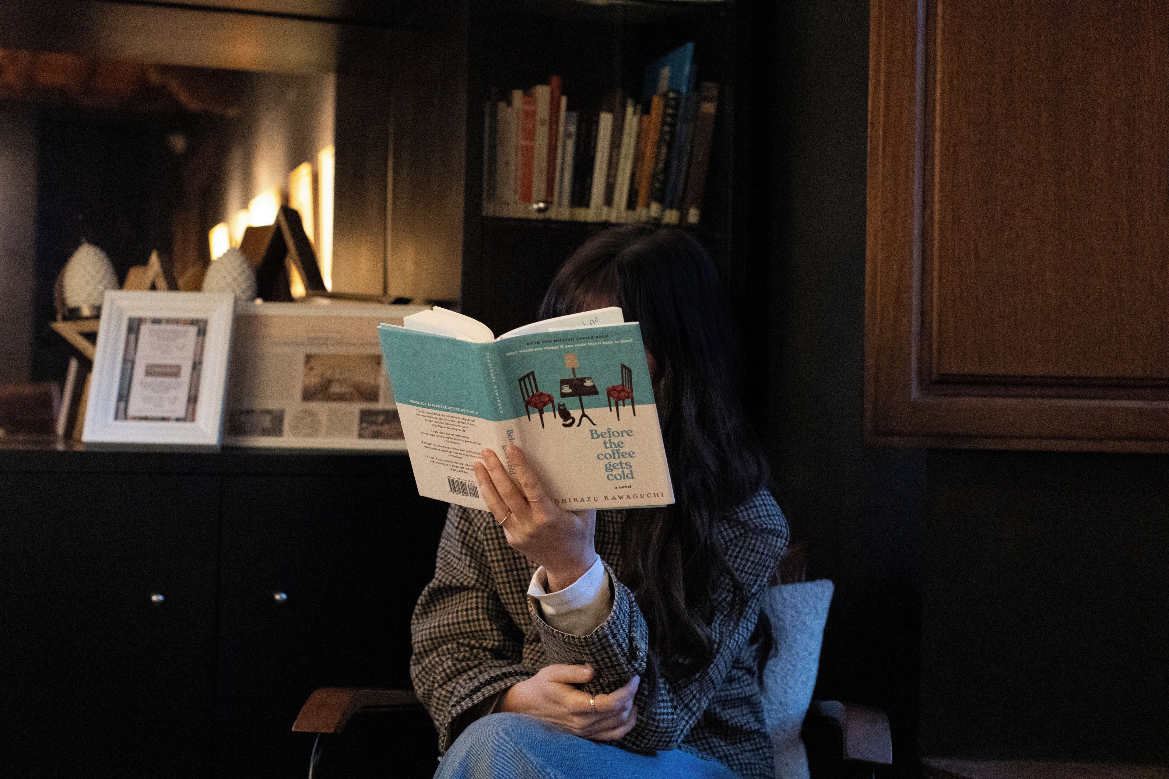 Uma pessoa lendo um livro em um quarto com estante e um quadro em cima de uma mesa.