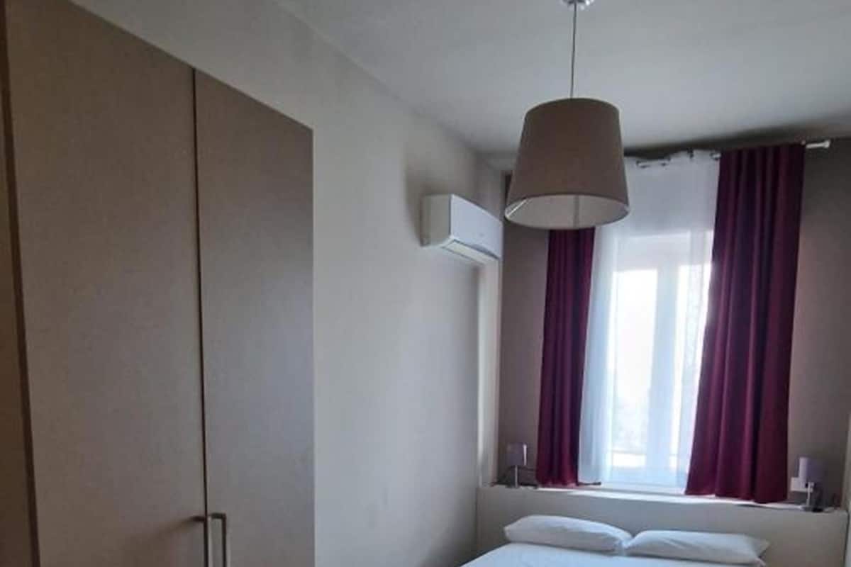 Quarto com cama, guarda-roupa, mesa de cabeceira, luminária pendente e janela com cortinas.
