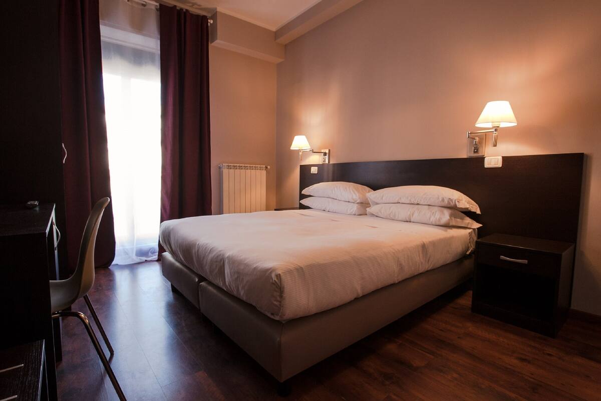 Quarto de hotel com cama, mesas de cabeceira, escrivaninha e cadeira.