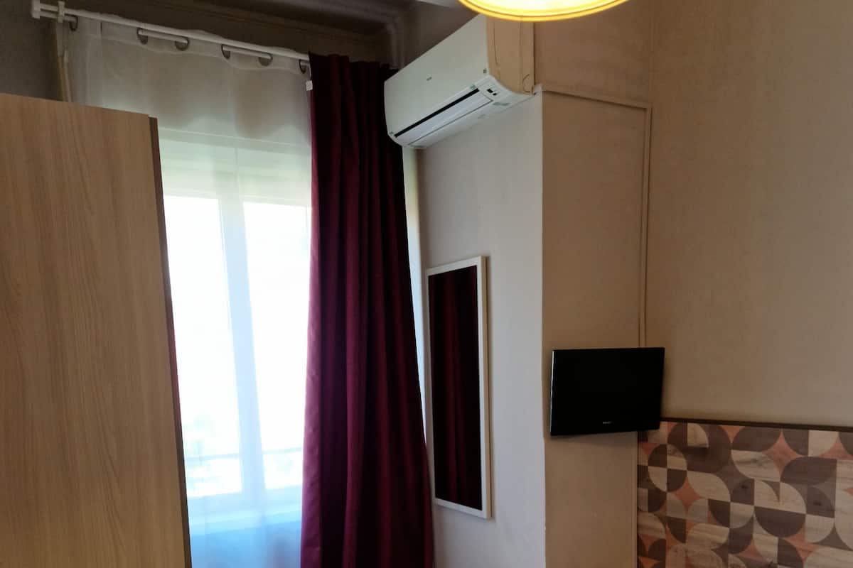Quarto pequeno com uma cama de solteiro, uma escrivaninha, uma cadeira, uma TV fixada na parede e aparelho de ar condicionado.