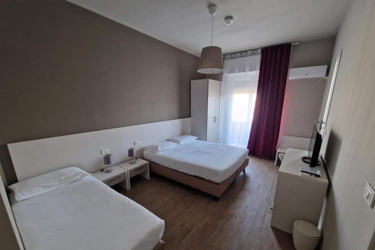 Quarto de hotel com duas camas, uma escrivaninha, uma cadeira, um abajur, um espelho e uma janela com cortinas.