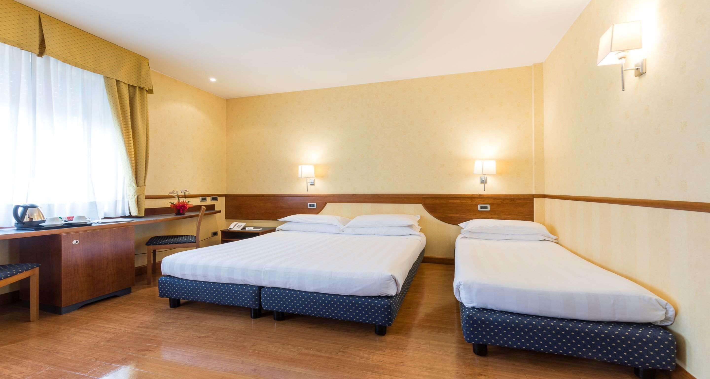 Quarto de hotel com duas camas, uma mesa de escritório, uma cadeira e uma pequena mesa com abajur.