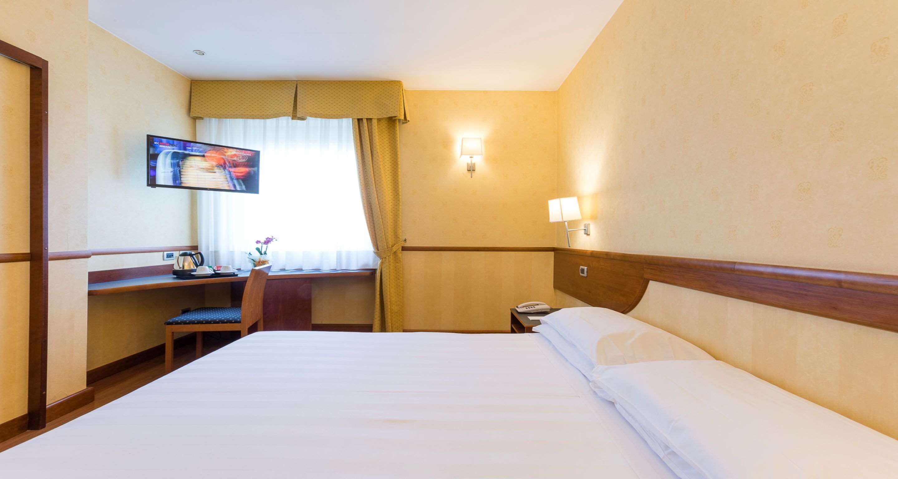 Quarto de hotel com cama, escrivaninha, cadeira, televisão e mesa de cabeceira com abajur.