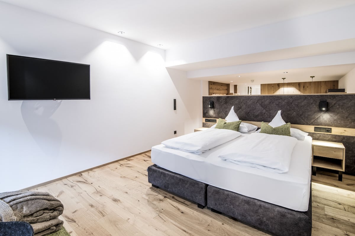 Quarto de hotel moderno com uma cama grande, uma TV de tela plana na parede, piso de madeira e uma área de cozinha compacta.