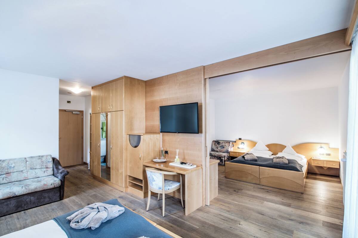Quarto de hotel moderno com uma escrivaninha de madeira, uma cama com cabeceira, um sofá e uma TV fixada na parede.