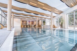 Uma piscina interior ampla com paredes de vidro transparente, vigas de madeira no teto e grandes janelas com vista para os arredores.