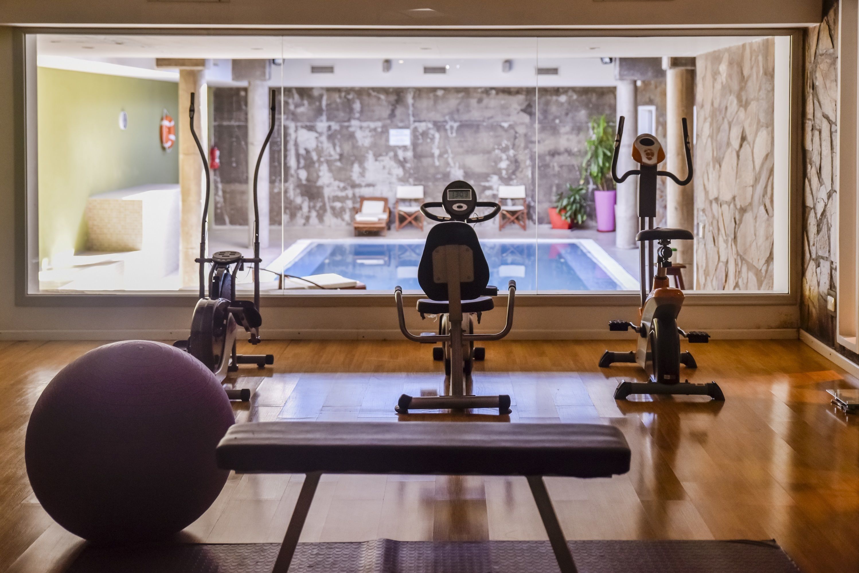 Academia com equipamentos de ginástica, uma bola de exercícios grande e vista para uma área de piscina externa.