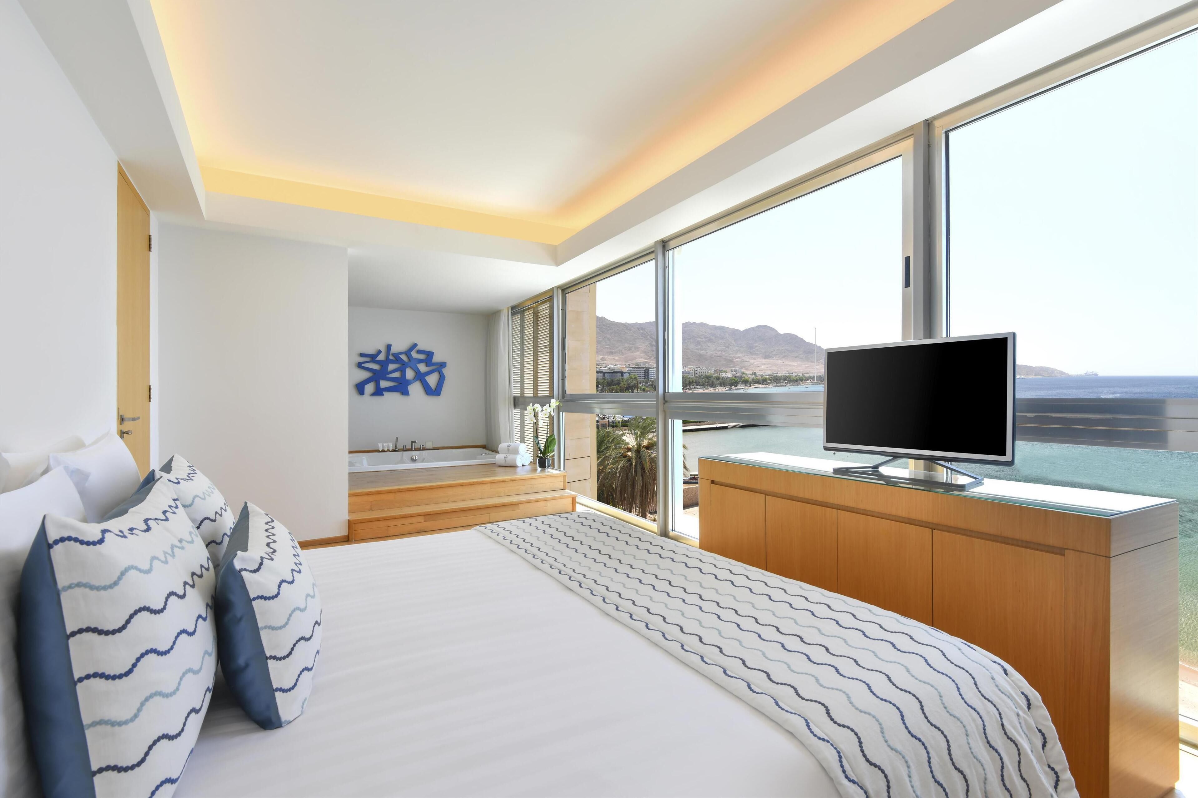 Quarto moderno com uma cama grande, uma TV em um console de madeira e vista para o mar.