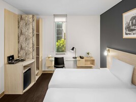 Quarto de hotel com cama, escrivaninha, cadeira e vista para a paisagem urbana pela janela.