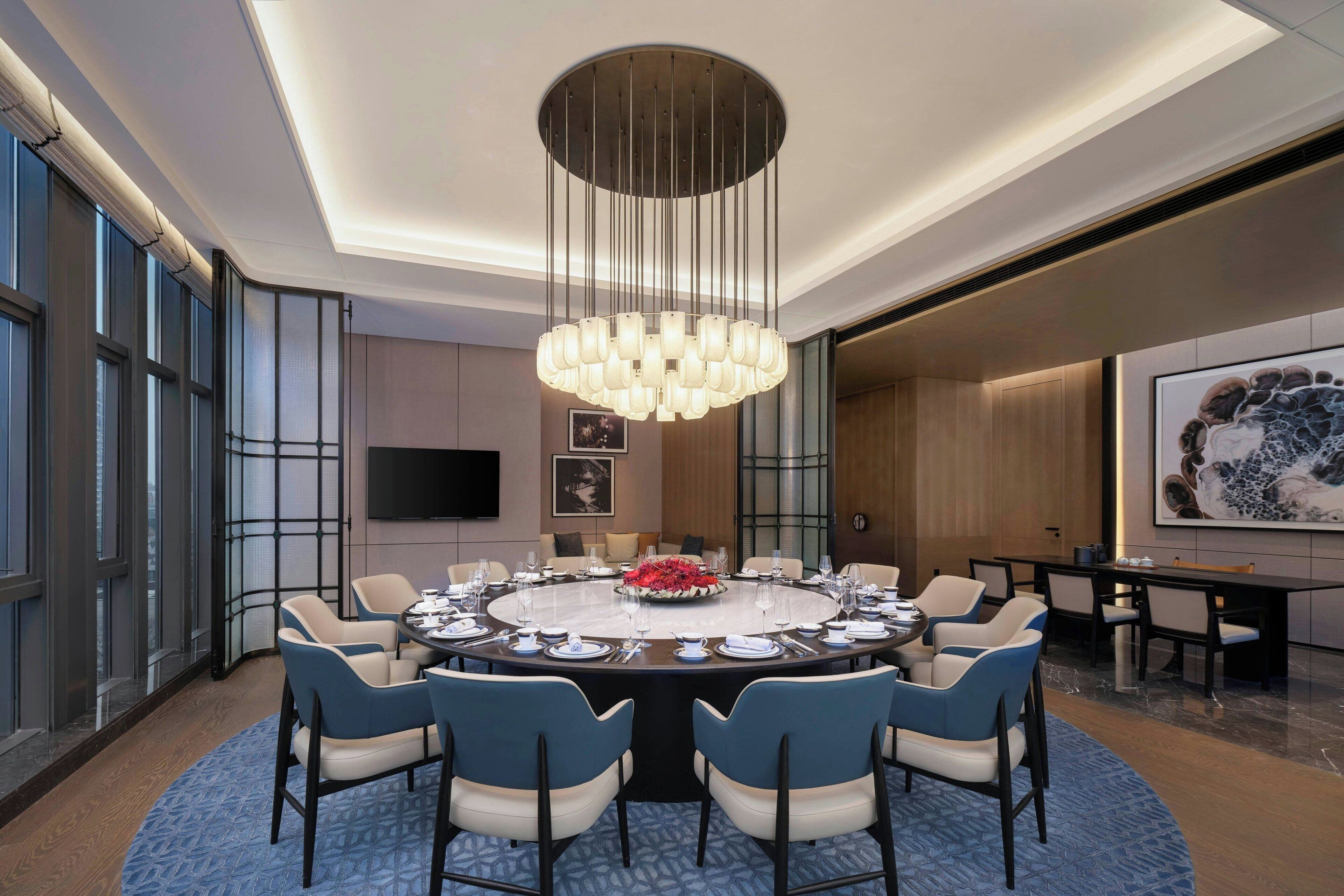 Uma sala de conferências moderna com uma mesa redonda, cadeiras azuis e bege, um candelabro e uma grande pintura abstrata na parede.