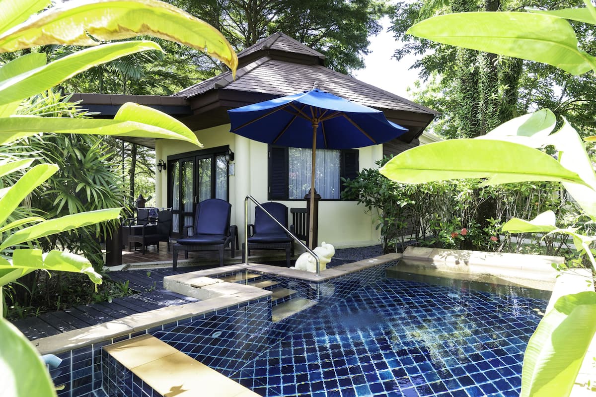 Uma área de piscina com piso em mosaico, um espaço coberto com cadeiras e um pequeno edifício com telhado inclinado.