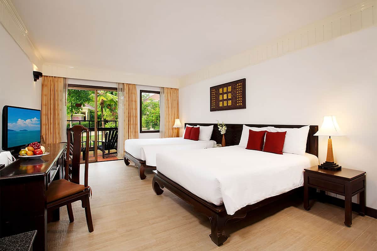 Quarto de hotel com duas camas, uma mesa com computador e vista para área verde lá fora.