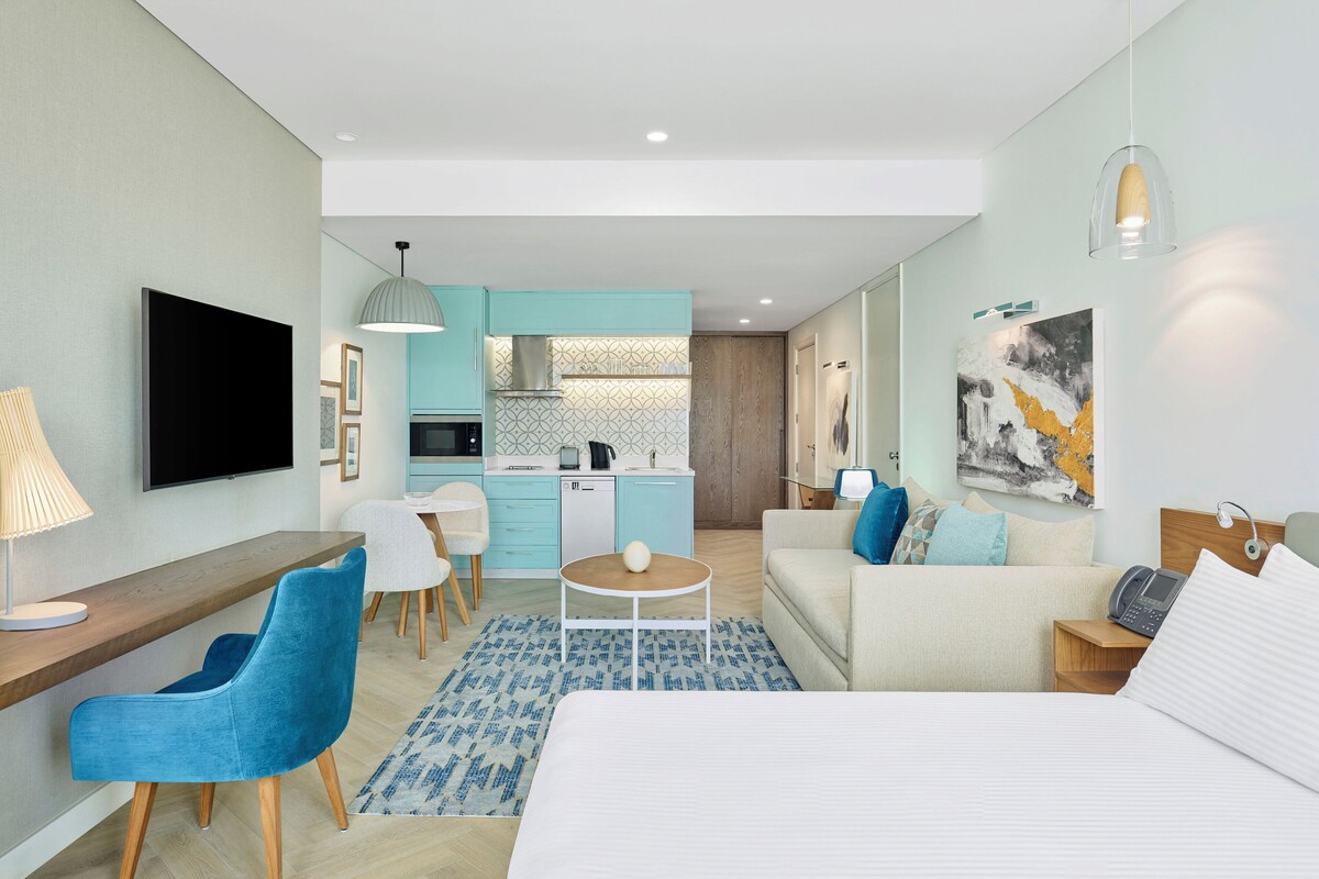 Quarto de hotel moderno com cama, escrivaninha com abajur, cadeira azul, mesinha redonda com uma bola, sofá com almofadas, área de cozinha ao fundo e um quadro na parede.