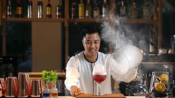 Um bar com um bartender preparando uma bebida, diversos copos e uma estante com garrafas ao fundo.