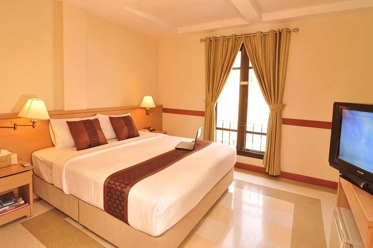 Quarto de hotel com cama, escrivaninha com luminária, televisão e janela com cortinas.