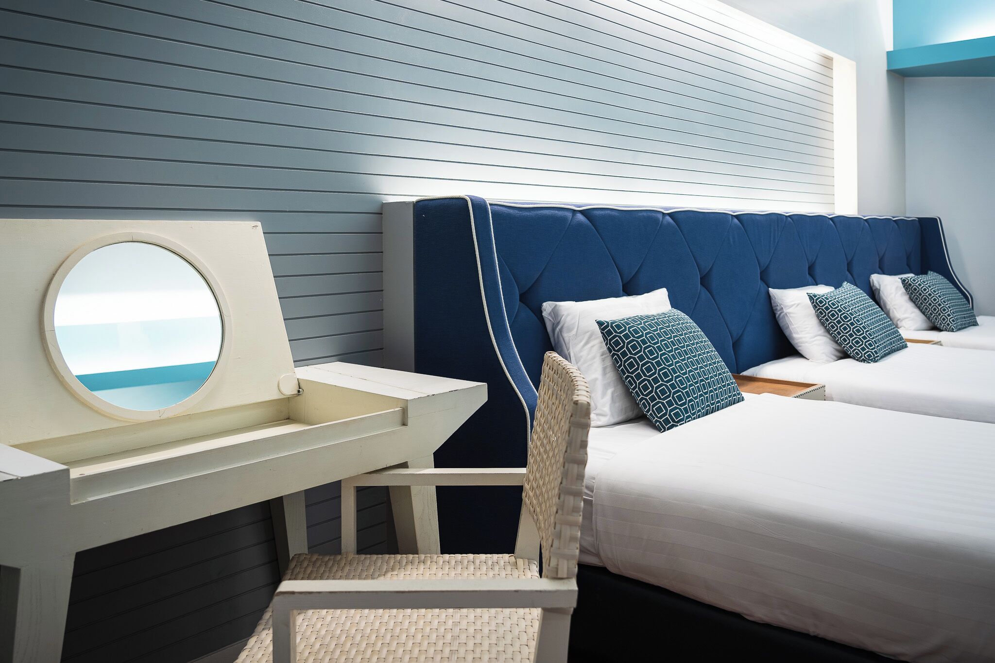 Quarto de hotel com uma janela grande, um espelho redondo, uma cama com roupa de cama branca e azul, e uma mesinha.