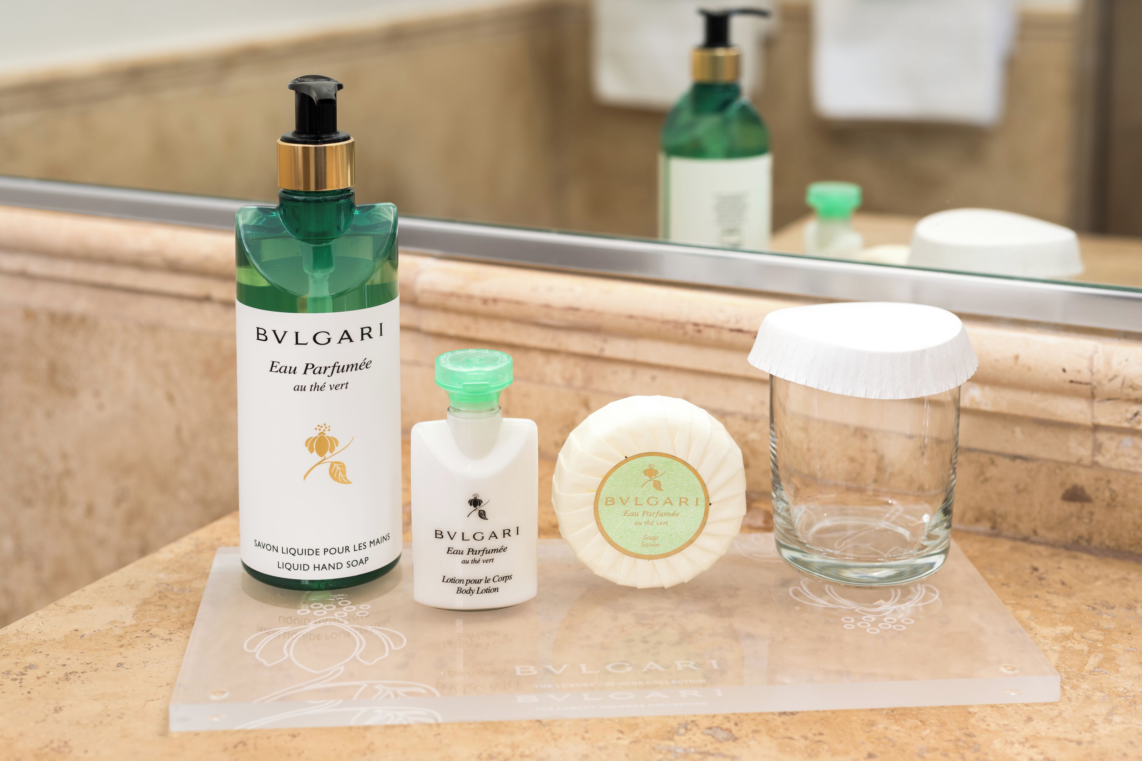 Uma bancada de banheiro com três frascos de Eau de Parfum Bvlgari, um frasco de loção corporal Bvlgari e um copo de água.