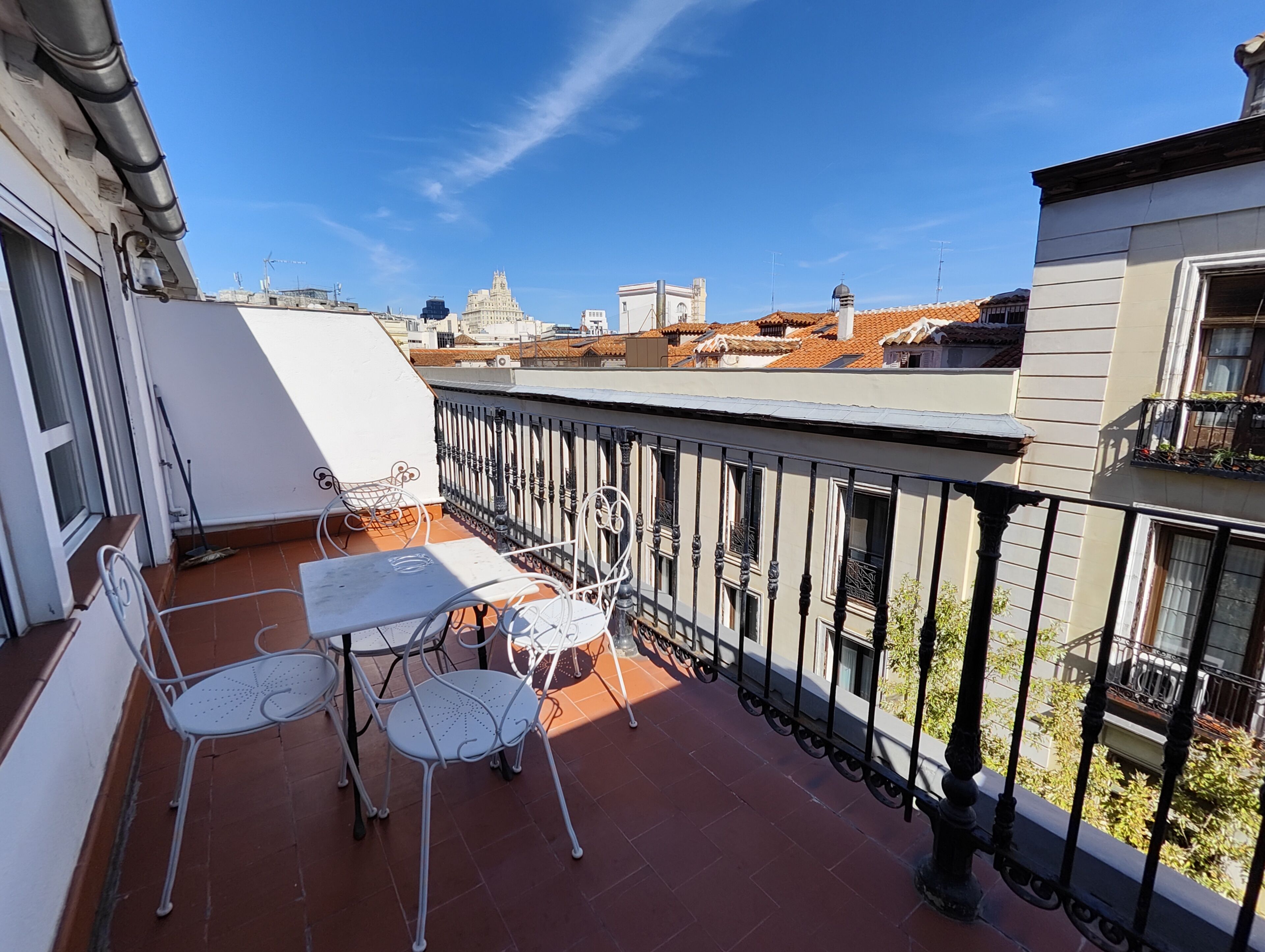 Uma varanda com mesa e cadeiras, grade branca e vista para edifícios ao longe.