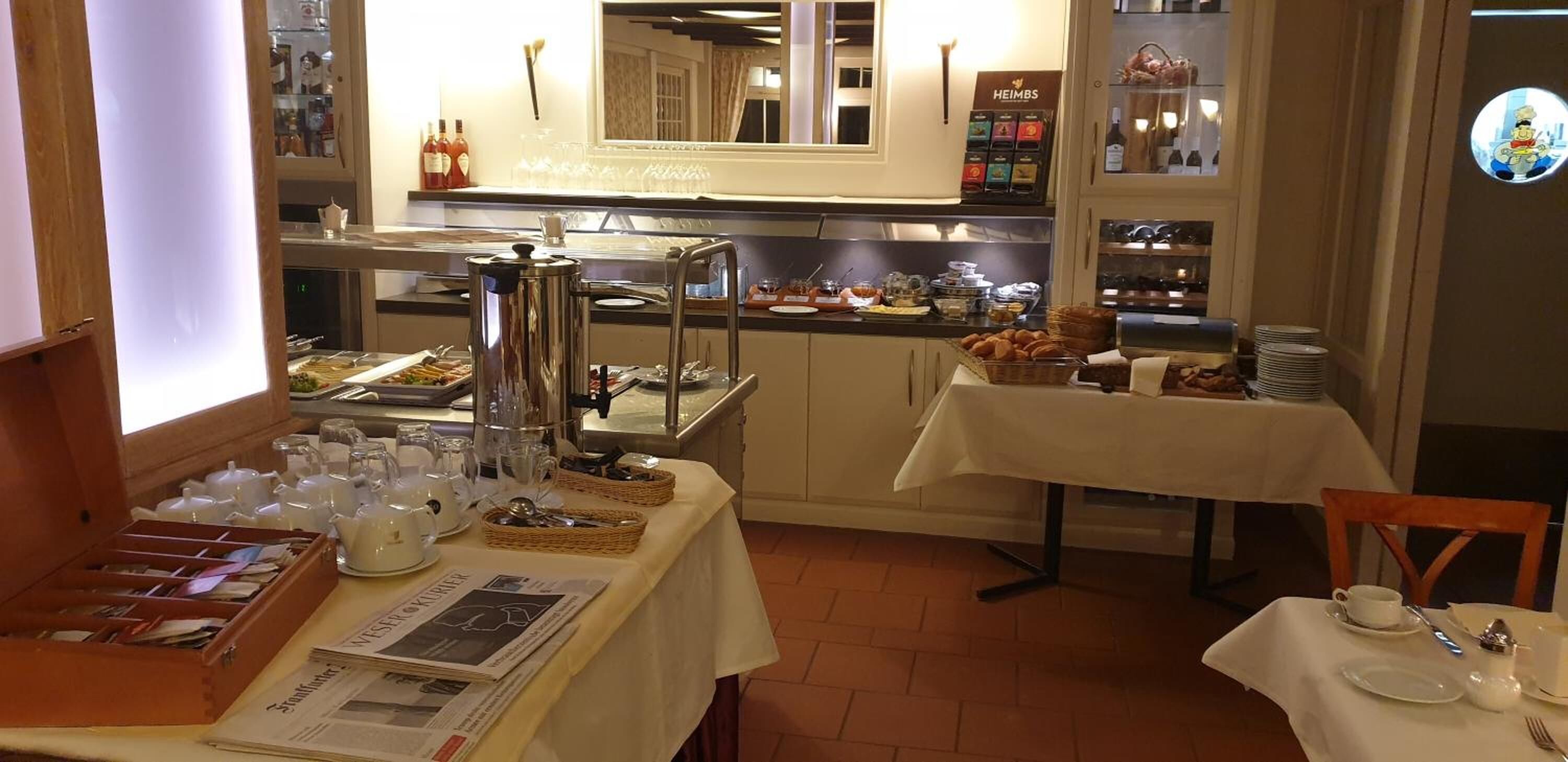 Um buffet de café da manhã com diversos alimentos e bebidas, uma máquina de café e um jornal sobre a mesa.