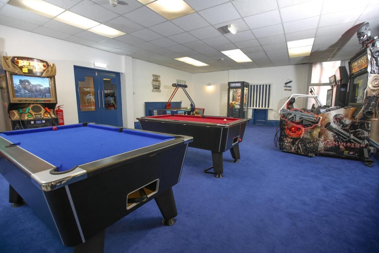 Uma mesa de bilhar com superfície azul, outra mesa de bilhar com superfície vermelha e uma máquina de garras ao fundo.