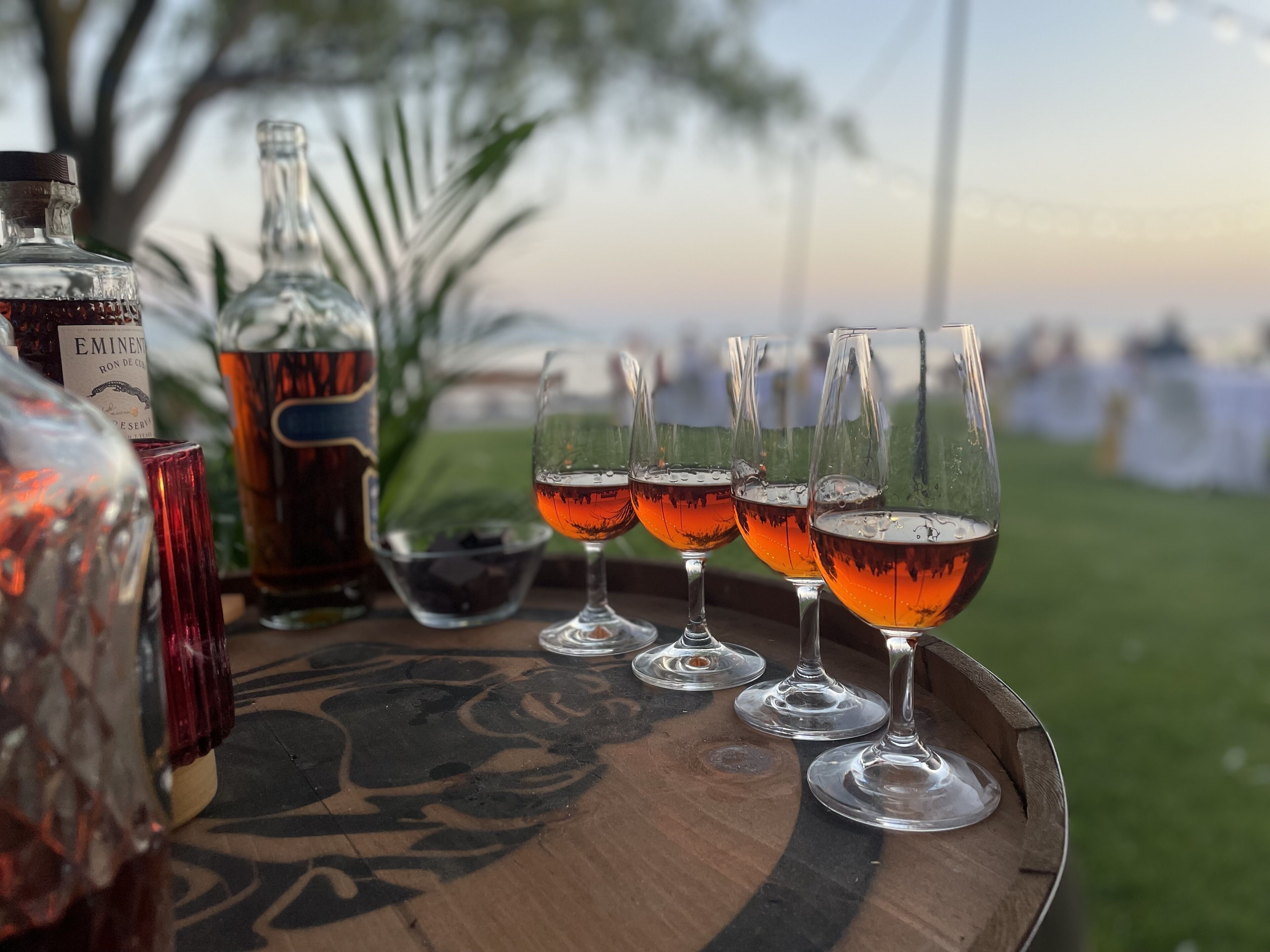 Uma mesa de madeira com quatro copos de vinho cheios de uma bebida avermelhada, uma garrafa de vinho Eminet e um recipiente de vidro com líquido vermelho.