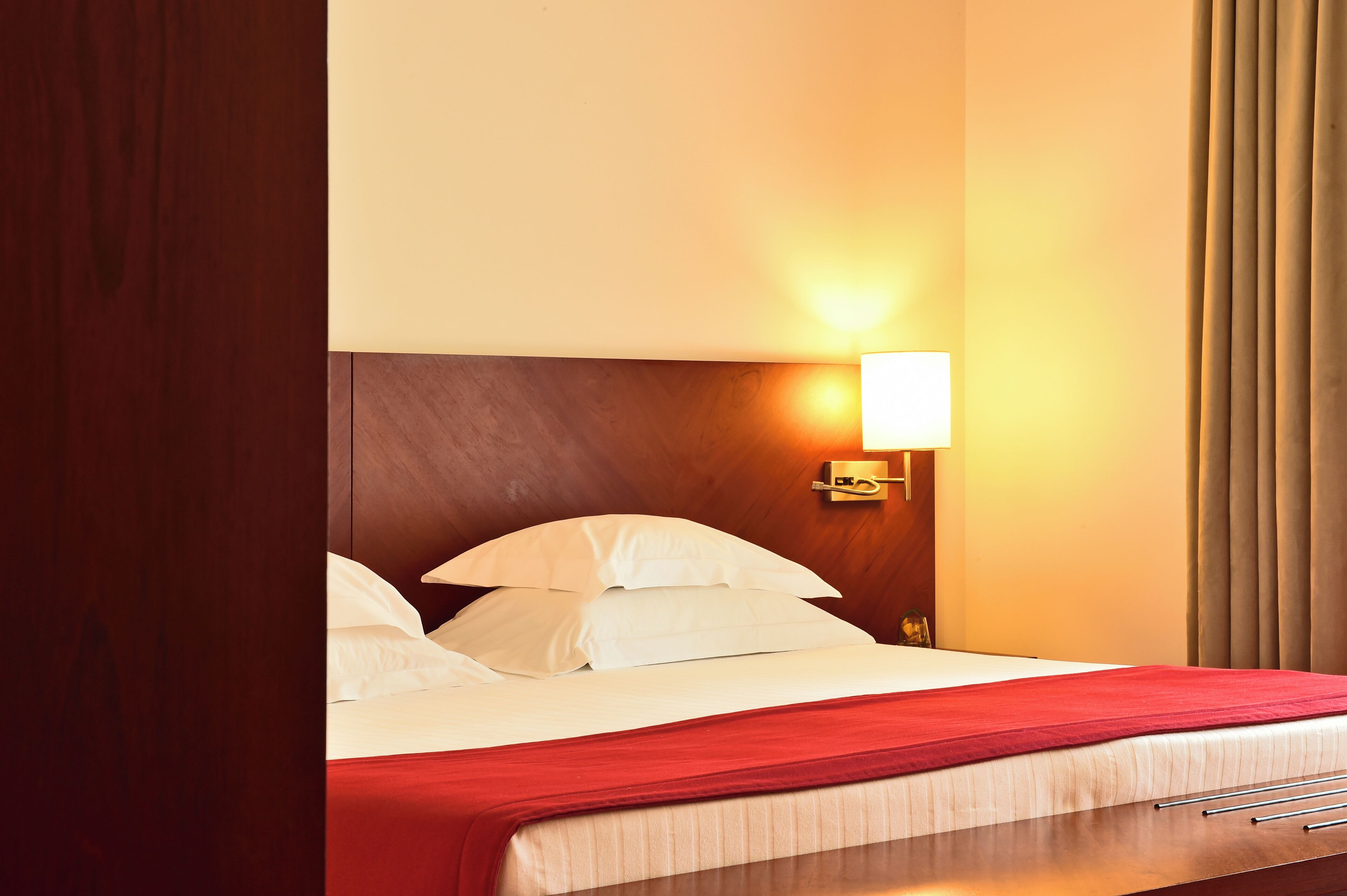 Quarto de hotel com cabeceira de madeira, cama com lençóis brancos, manta vermelha e luminária fixa na parede.