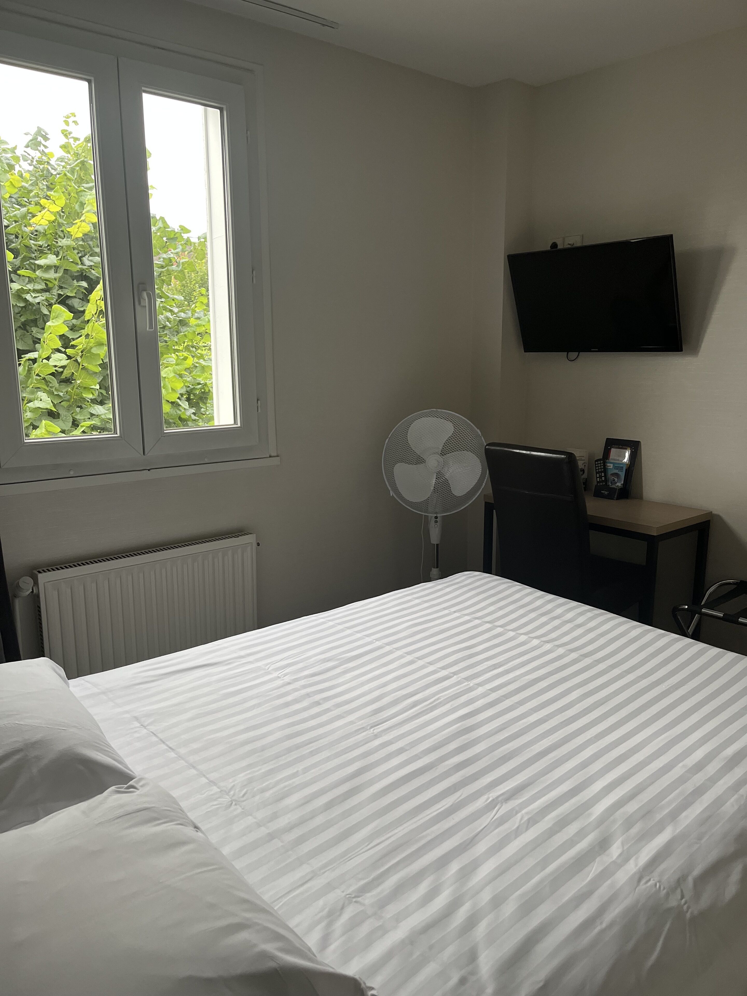 Quarto de hotel com cama, escrivaninha com computador, televisão e janela com vista para áreas verdes.