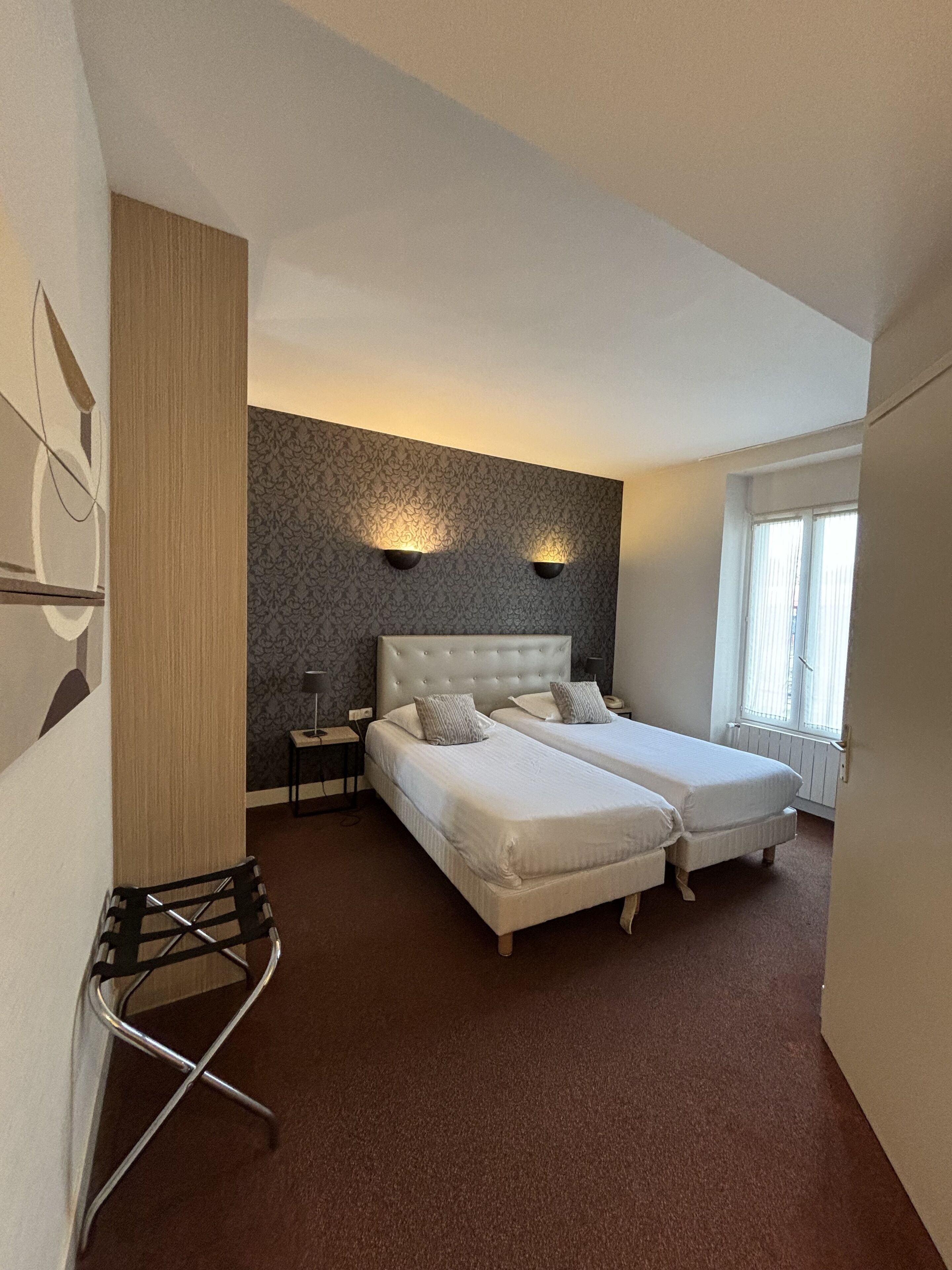 Quarto de hotel com duas camas, uma mesinha, uma cadeira e uma janela.