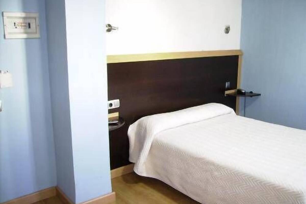 Quarto pequeno com uma cama de solteiro, cabeceira de madeira, mesa de cabeceira e luminária fixada na parede.