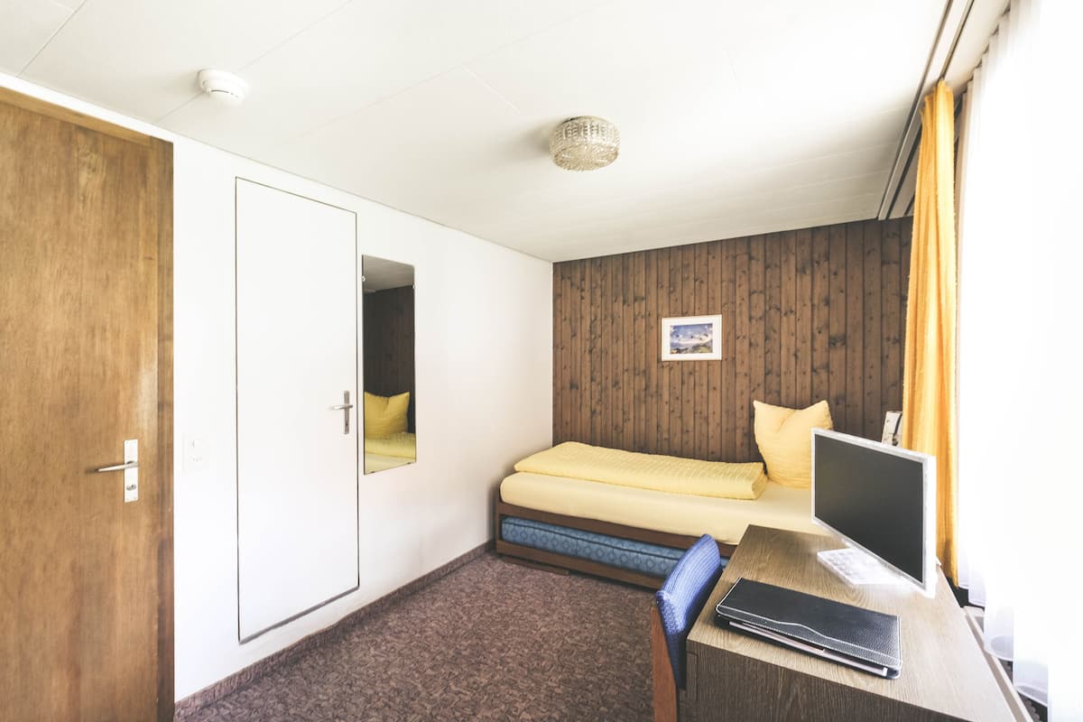 Quarto de hotel pequeno com uma cama de solteiro, uma escrivaninha com computador, um espelho e parede de painel de madeira.