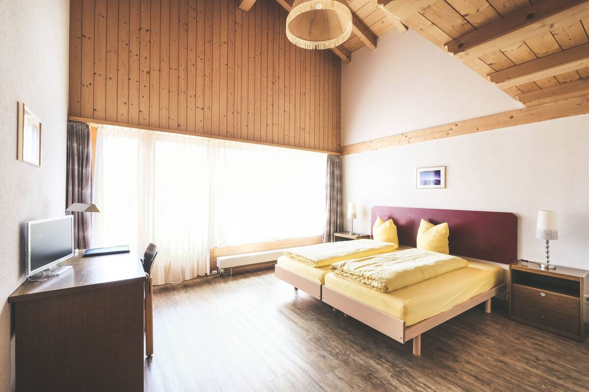 Quarto com teto de madeira, cama com roupa de cama amarela, escrivaninha com computador e janela com cortinas.