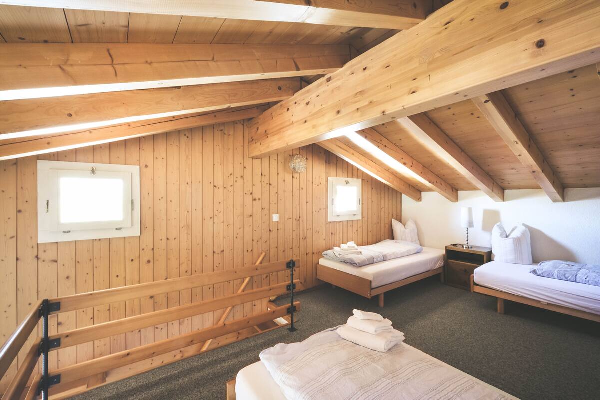 Quarto de sótão com paredes de madeira, duas camas, uma janela pequena e escada de madeira.
