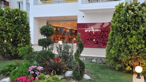Um edifício moderno com entrada de vidro, rodeado por jardins bem cuidados e um muro decorativo.