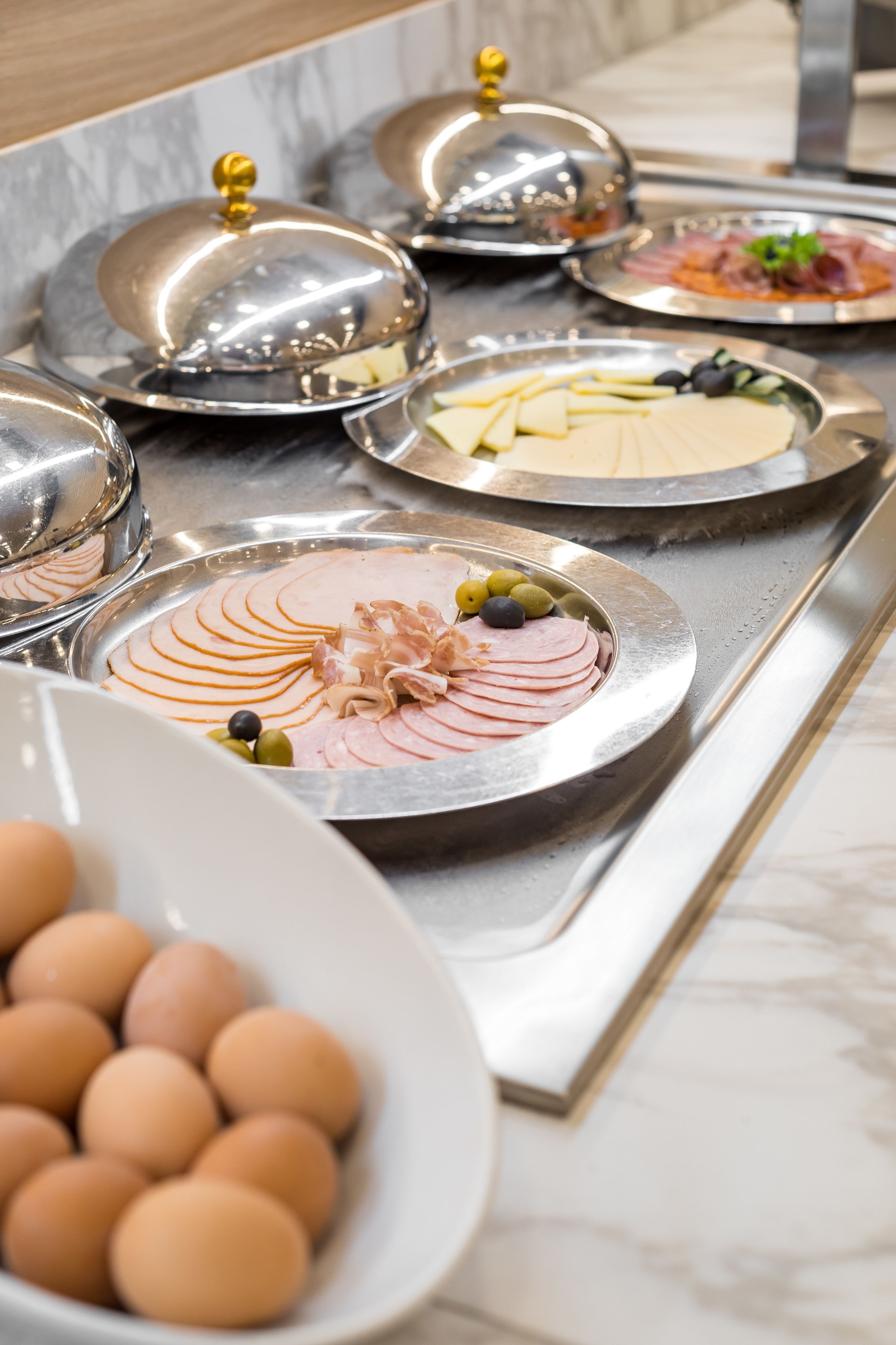 Buffet com diversos pratos, incluindo carnes e queijos fatiados, cobertos por cloches de prata.