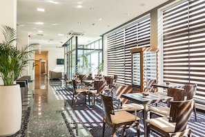Um saguão de hotel moderno com divisória de vidro, cadeiras de madeira e uma grande planta em vaso.