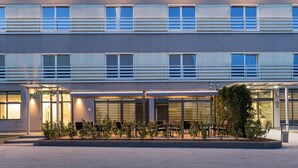 Um edifício moderno com restaurante no andar térreo, com mesas ao ar livre e entrada bem iluminada.