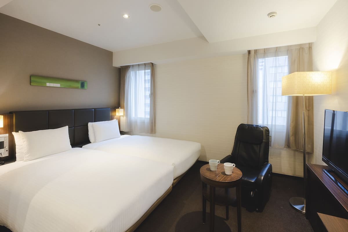 Quarto de hotel com uma cama grande, uma escrivaninha com duas chávenas, uma cadeira, uma televisão e um candeeiro.