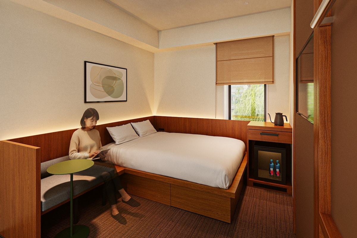 Quarto de hotel pequeno com cama, mesa, cadeira, mesa de cabeceira com abajur e janela com vista.