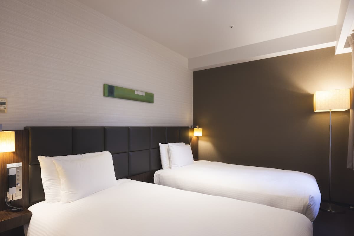 Quarto de hotel com duas camas, mesa de cabeceira com abajur, painel de controle na parede e uma janela grande.