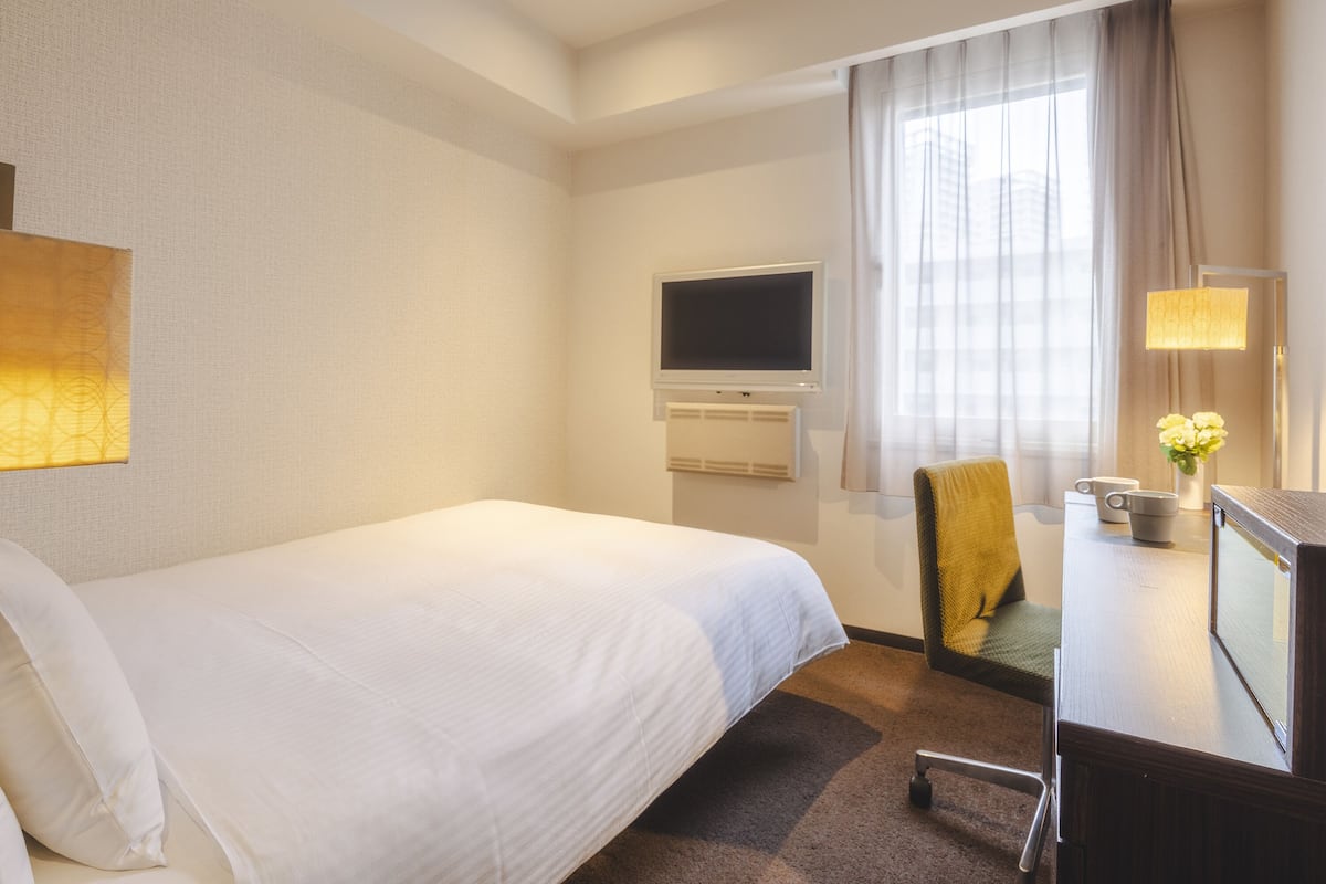 Quarto de hotel com uma cama, uma escrivaninha com abajur, uma televisão e uma janela com cortinas.