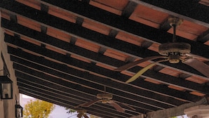 Uma área de estar coberta ao ar livre, com cadeiras e mesas brancas, colunas e vista para o exterior.